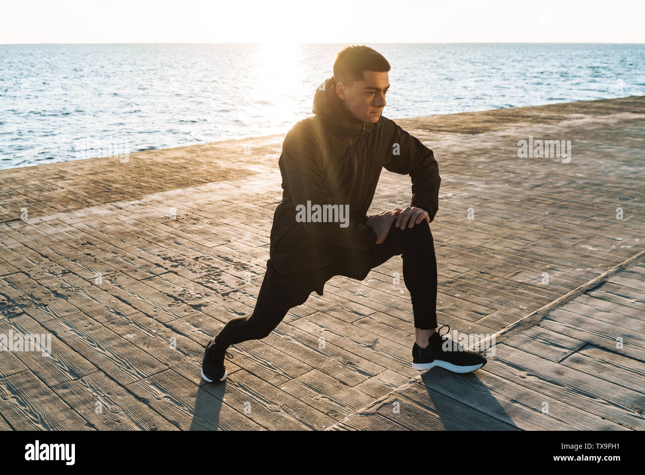 Confident young sportsman wearing jacket warming up at the beach, doing ...