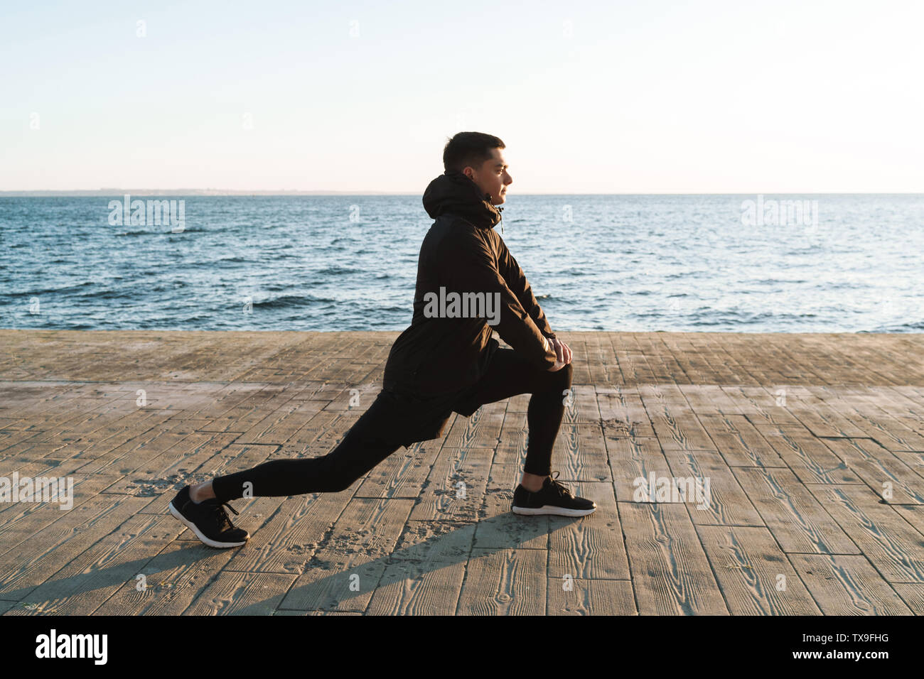 Confident young sportsman wearing jacket warming up at the beach, doing ...