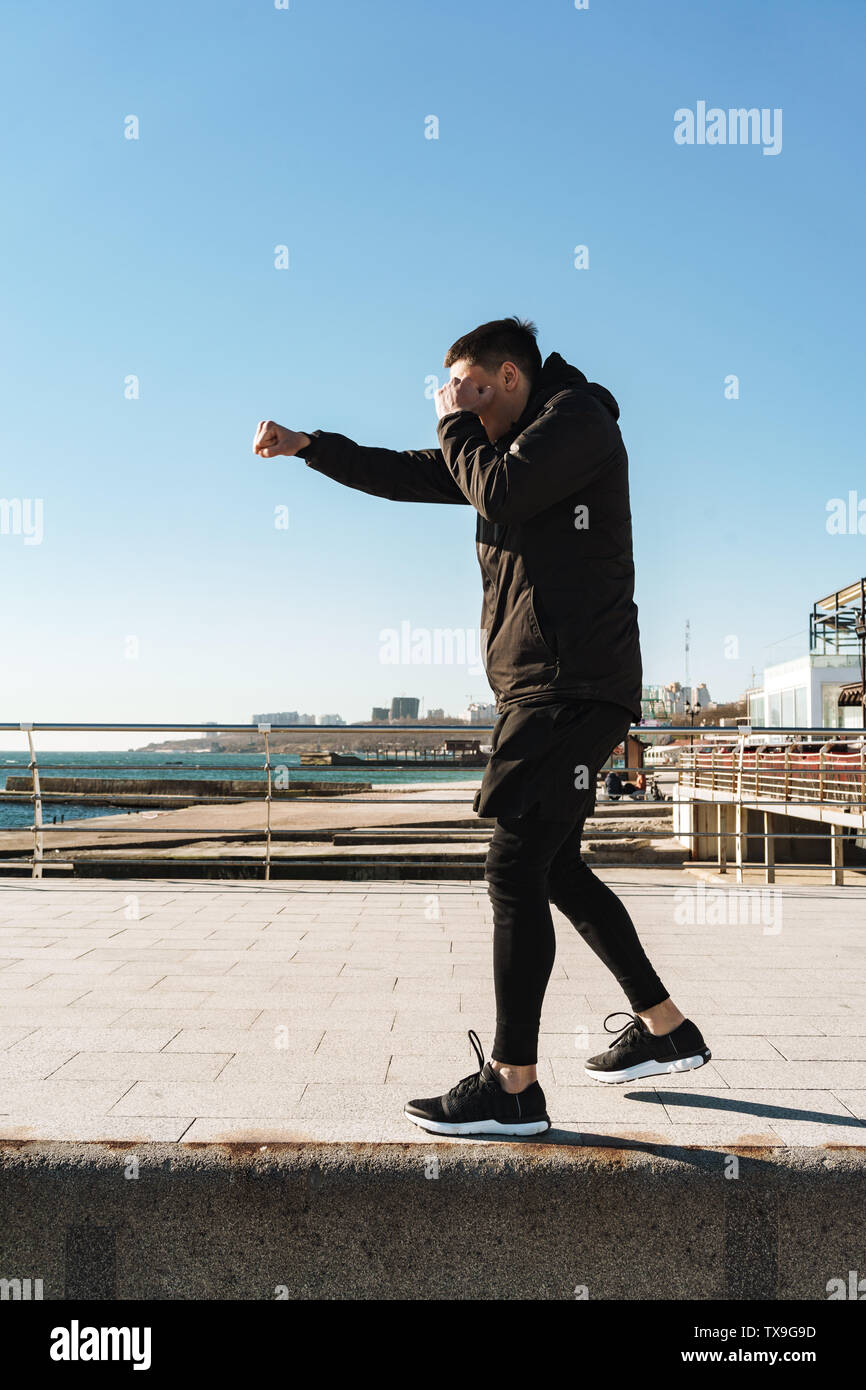 Photo of sporty man 20s in black tracksuit boxing and doing punch ...