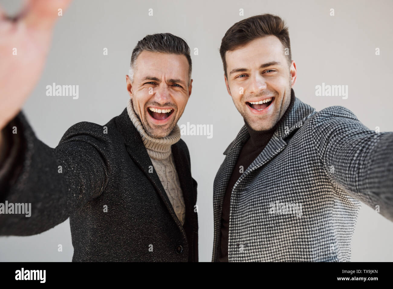 Two handsome men wearing coats over gray background, taking a selfie ...