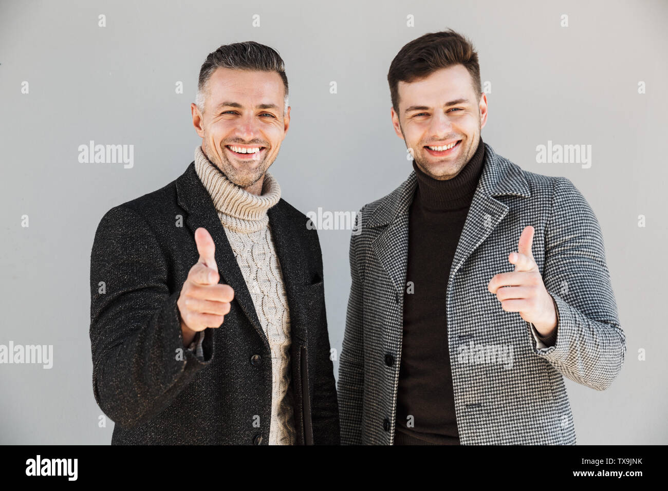 Two handsome men wearing coats standing over gray background, pointing ...