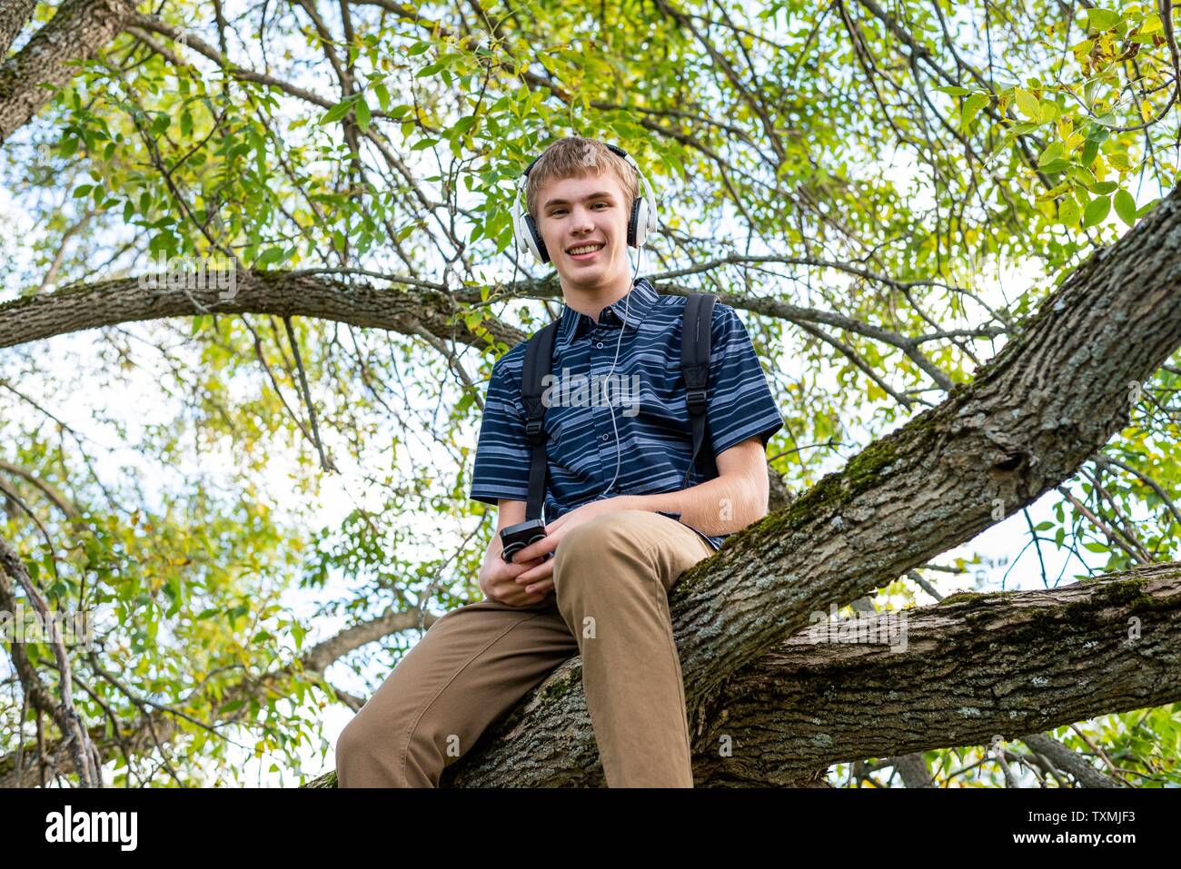 Happy student sitting on a tree branch while listening to music through ...