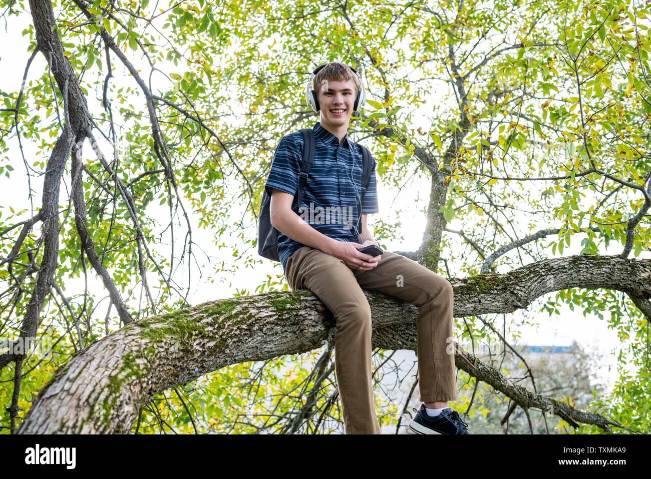 Happy student sitting on a tree branch while listening to music through ...