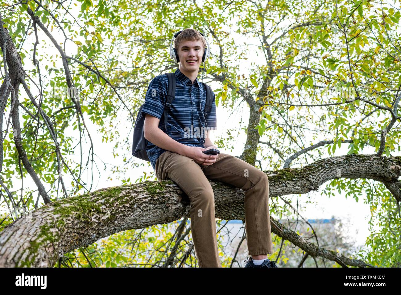 Happy student sitting on a tree branch while listening to music through ...
