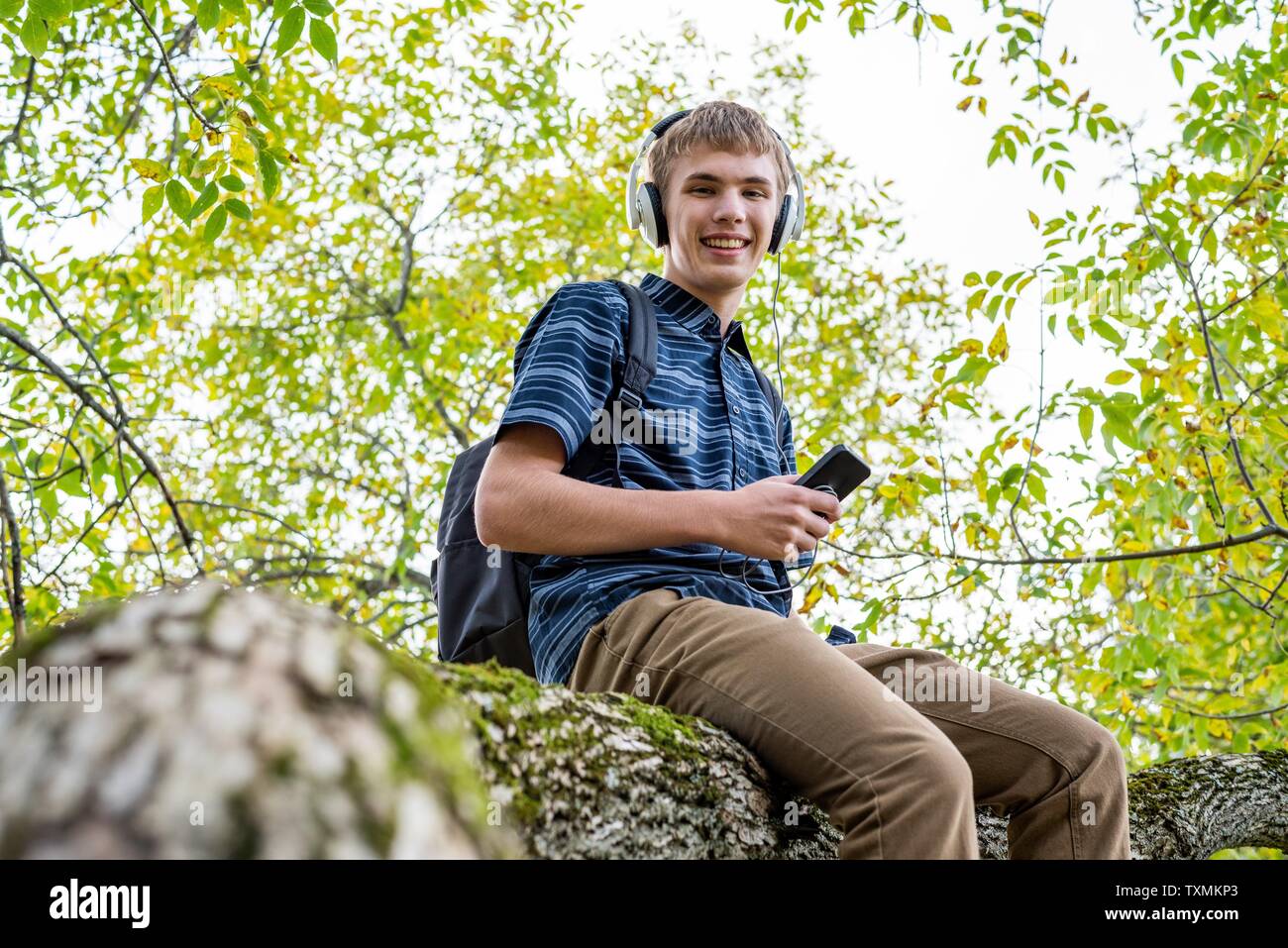 Happy student sitting on a tree branch while listening to music through ...