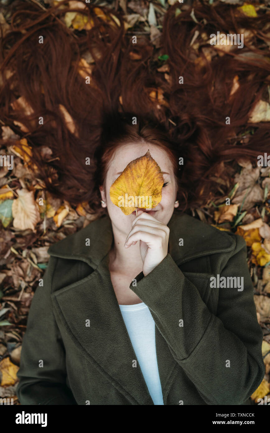 Young woman with long red hair lying amongst autumn leaves and covering ...
