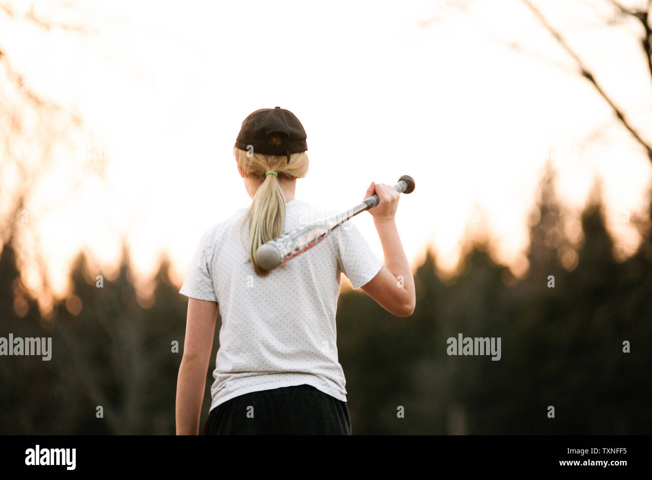 Girl with baseball bat over her shoulder in rural field, rear view ...