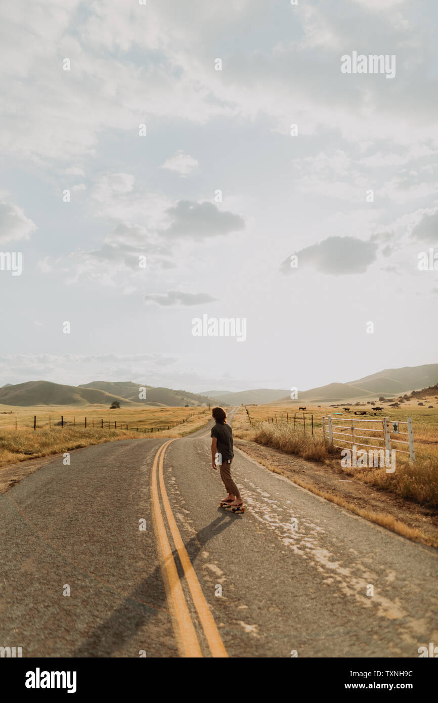 Young barefoot male skateboarder skateboarding on rural road, rear view ...