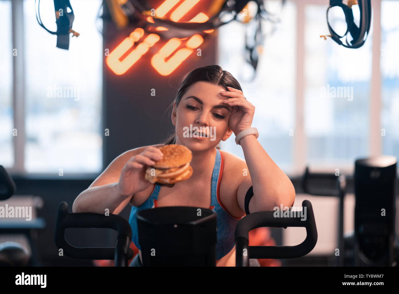 Woman looking at burger while sitting on exercise cycle Stock Photo - Alamy