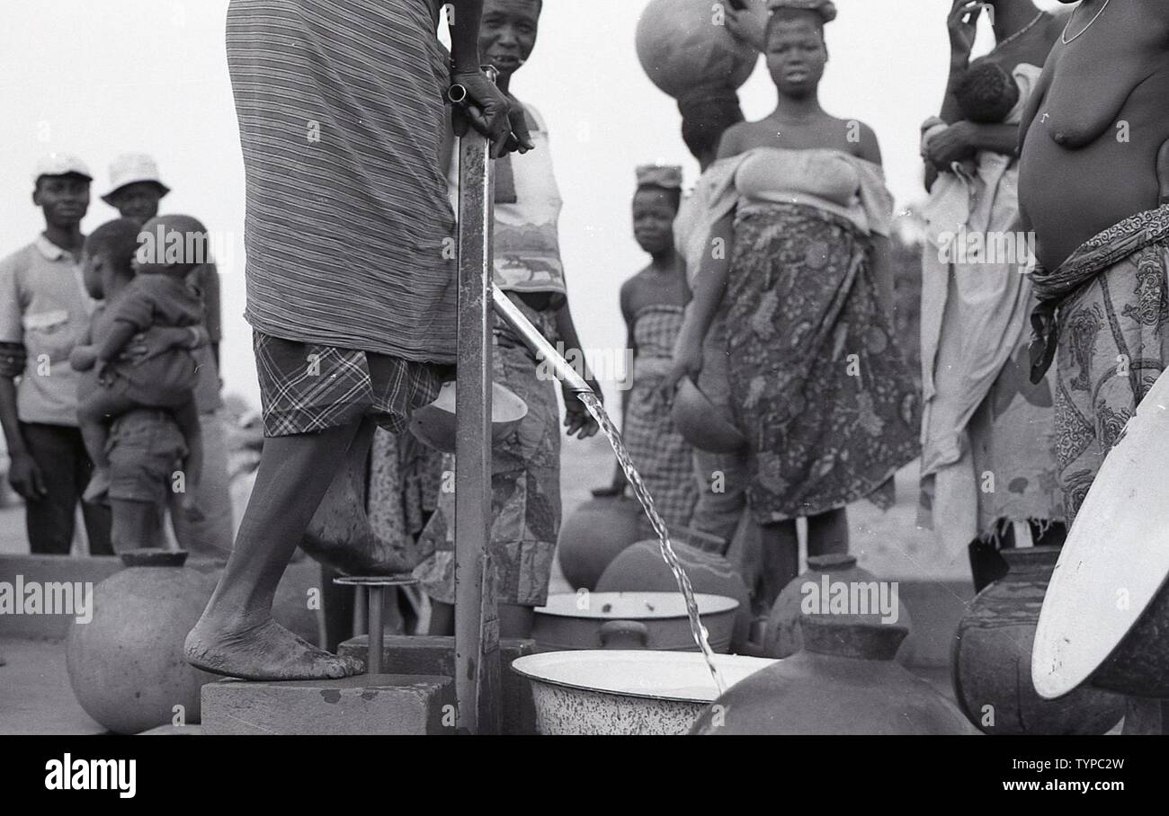 WOMAN PUMPING WATER FROM A WELL, WHILE OTHERS STAND AROUND WITH ...