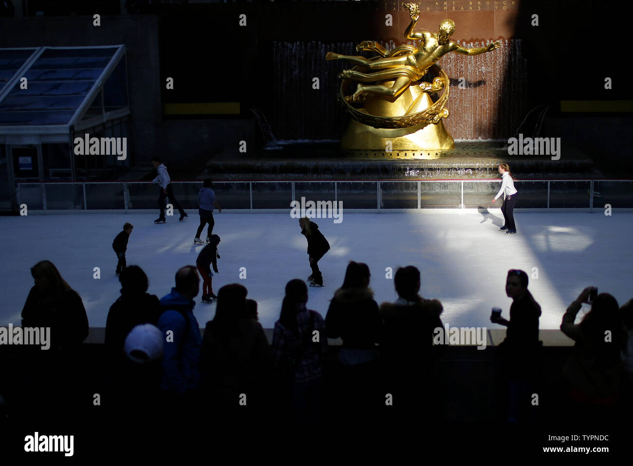 A crowd gathers to watch people ice skating below a gold statue of ...