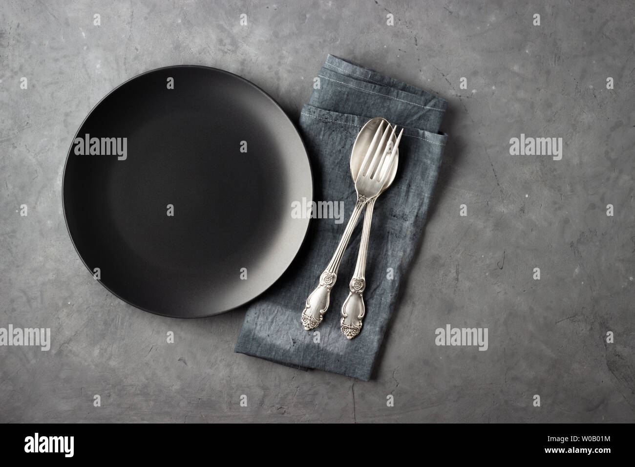 Table setting. Black plate, cutlery and napkin over gray concrete ...