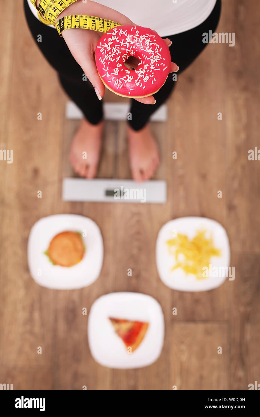 Young beautiful woman standing on scales and holding a donut. The ...