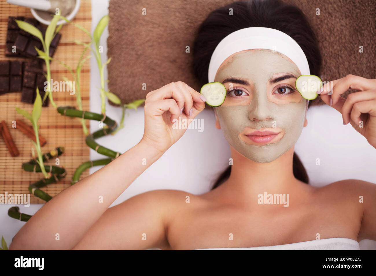 Spa. Beautiful young woman is getting facial clay mask at spa, lying with cucumbers on eyes ...