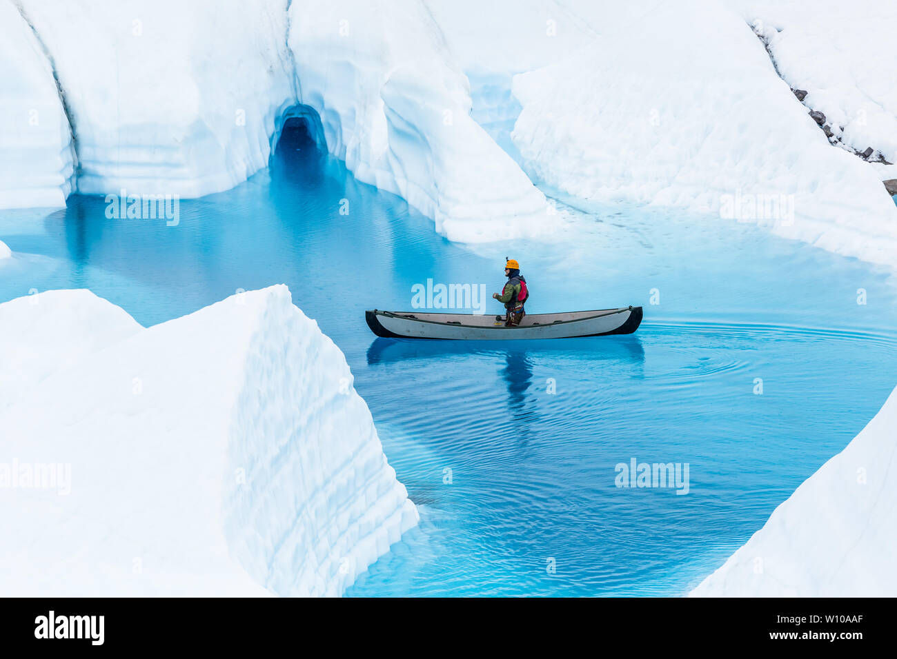 Man paddles a canoe in front of a small flooded ice cave on a glacier ...