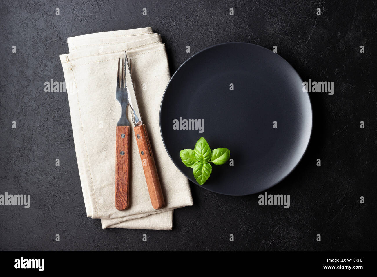 Table setting. Black plate, cutlery, napkin and basil leaves over black ...
