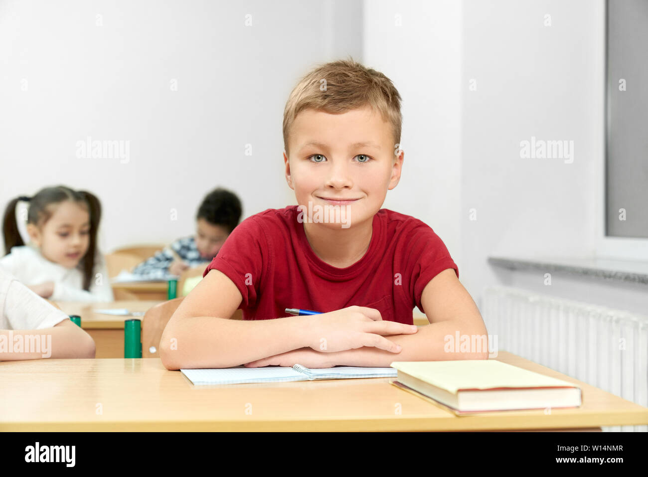 Front view of little male student sitting at desk with folded arms ...