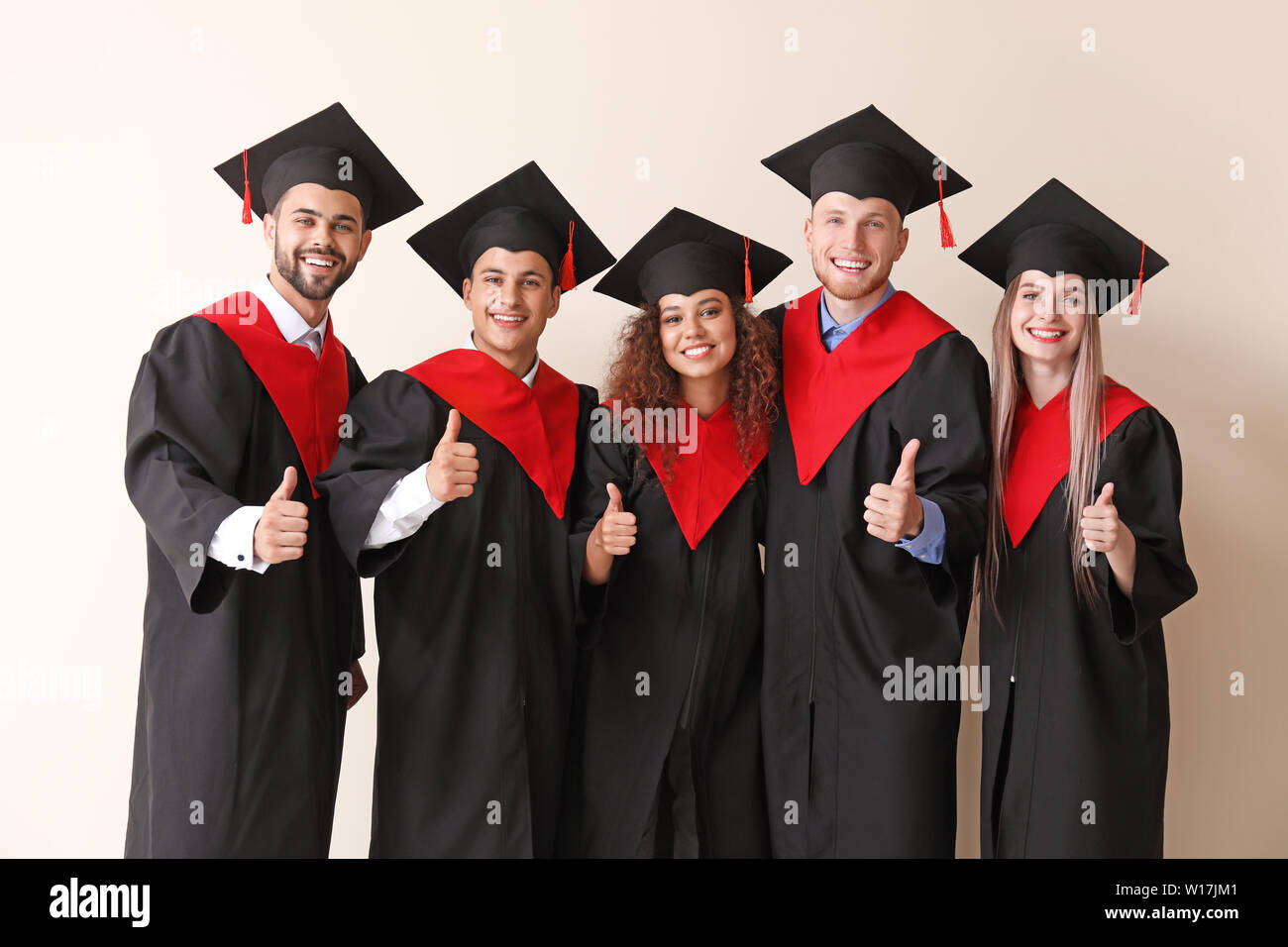 Young students in bachelor robes showing thumb-up on light background ...
