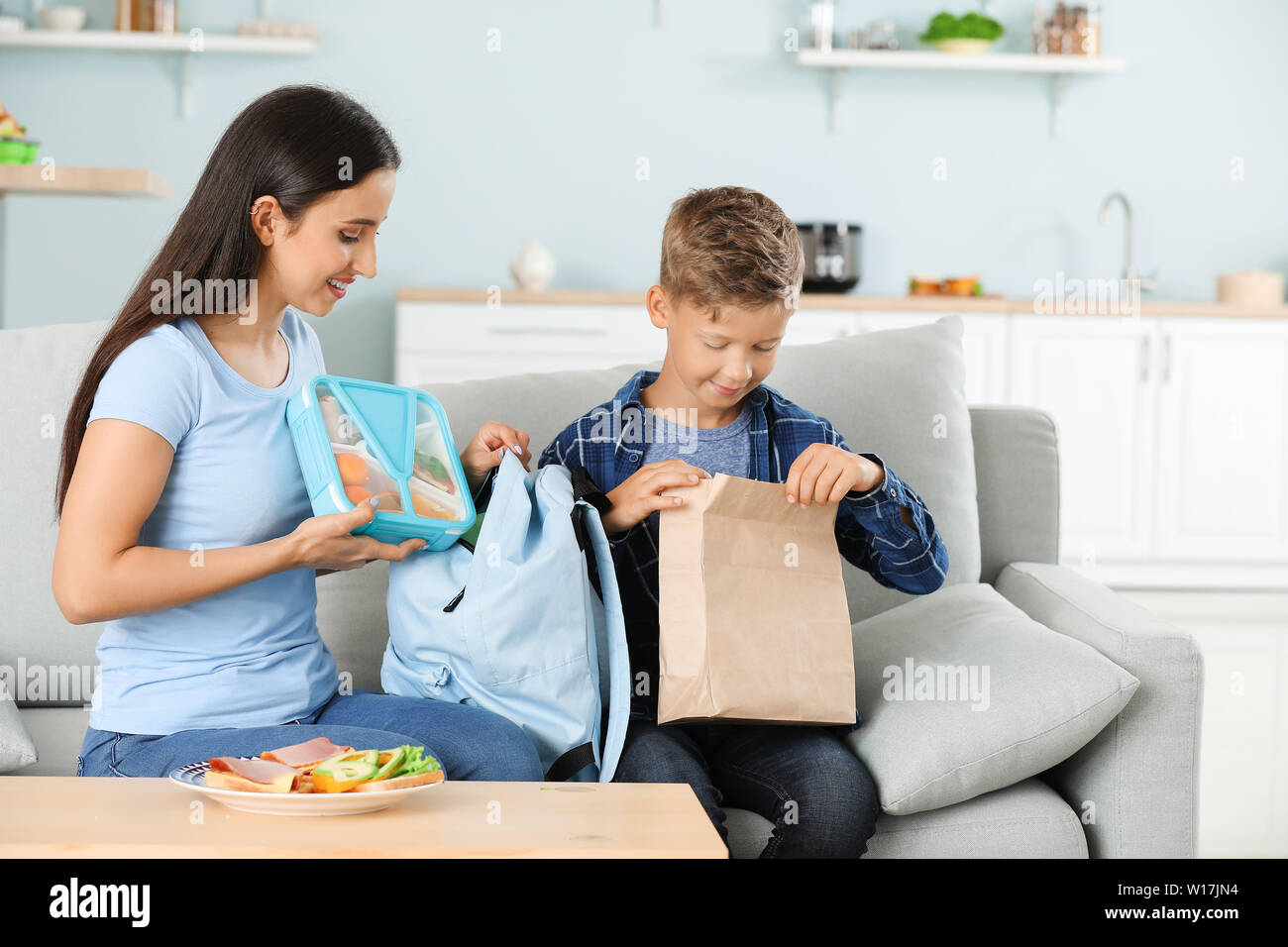 Mother putting lunch box into backpack of her little son before school ...
