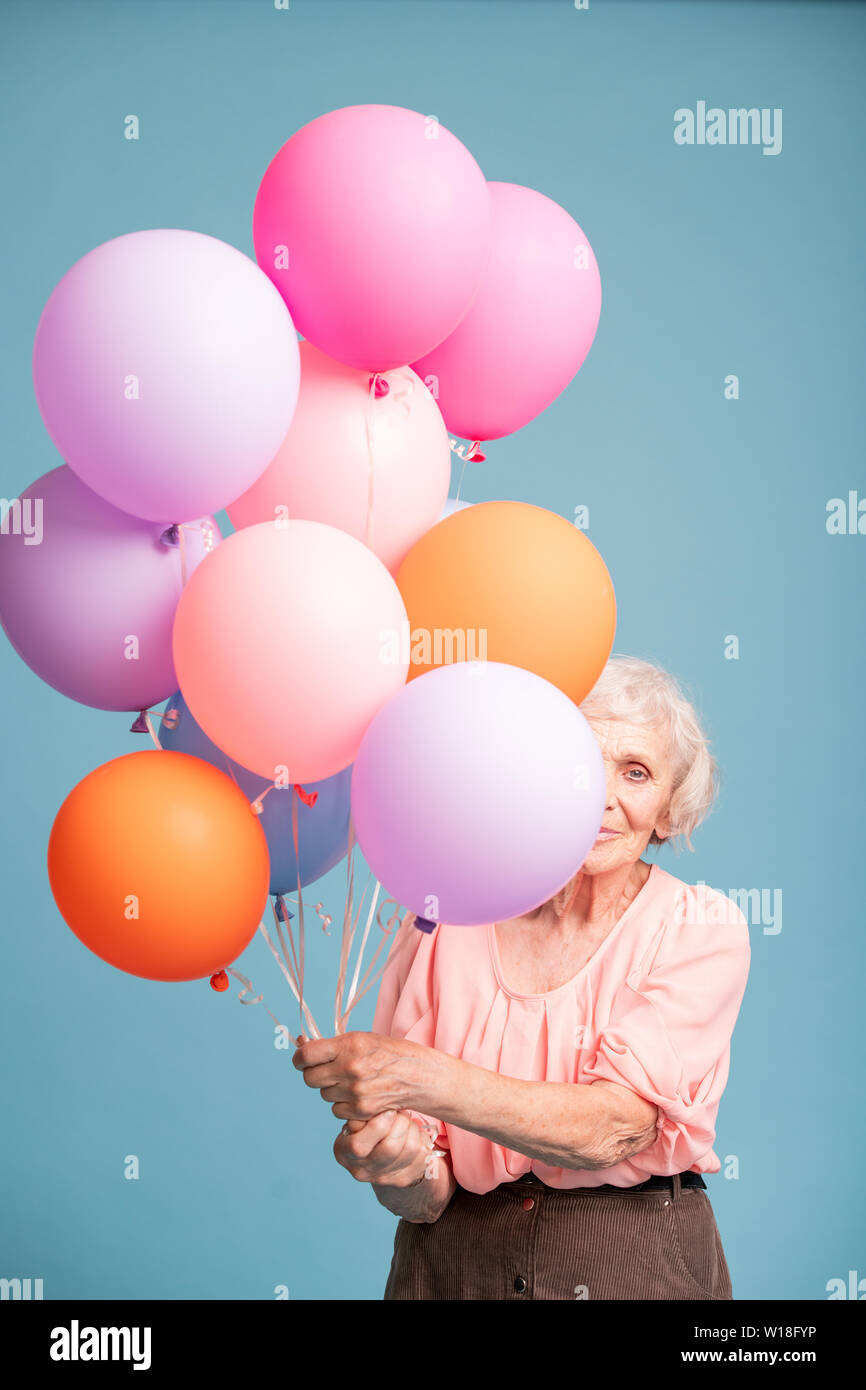 Happy senior woman peeking out of bunch of colorful balloons in ...