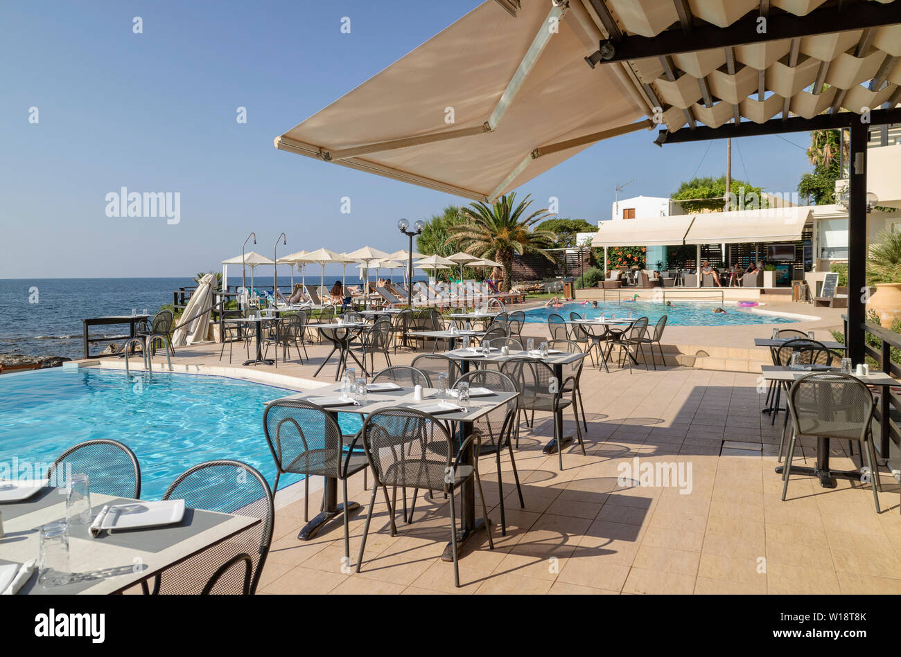 Crete, Greece. June 2019. Poolside restaurant table and chairs overlook ...