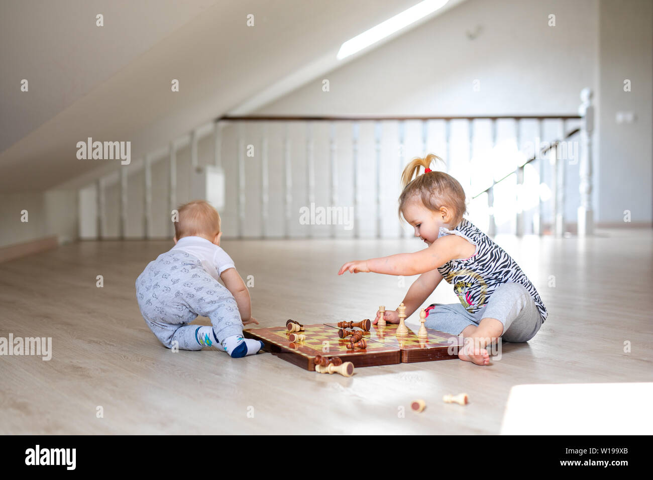 Two babies boy and girl playing chess on the white wooden floor at home ...
