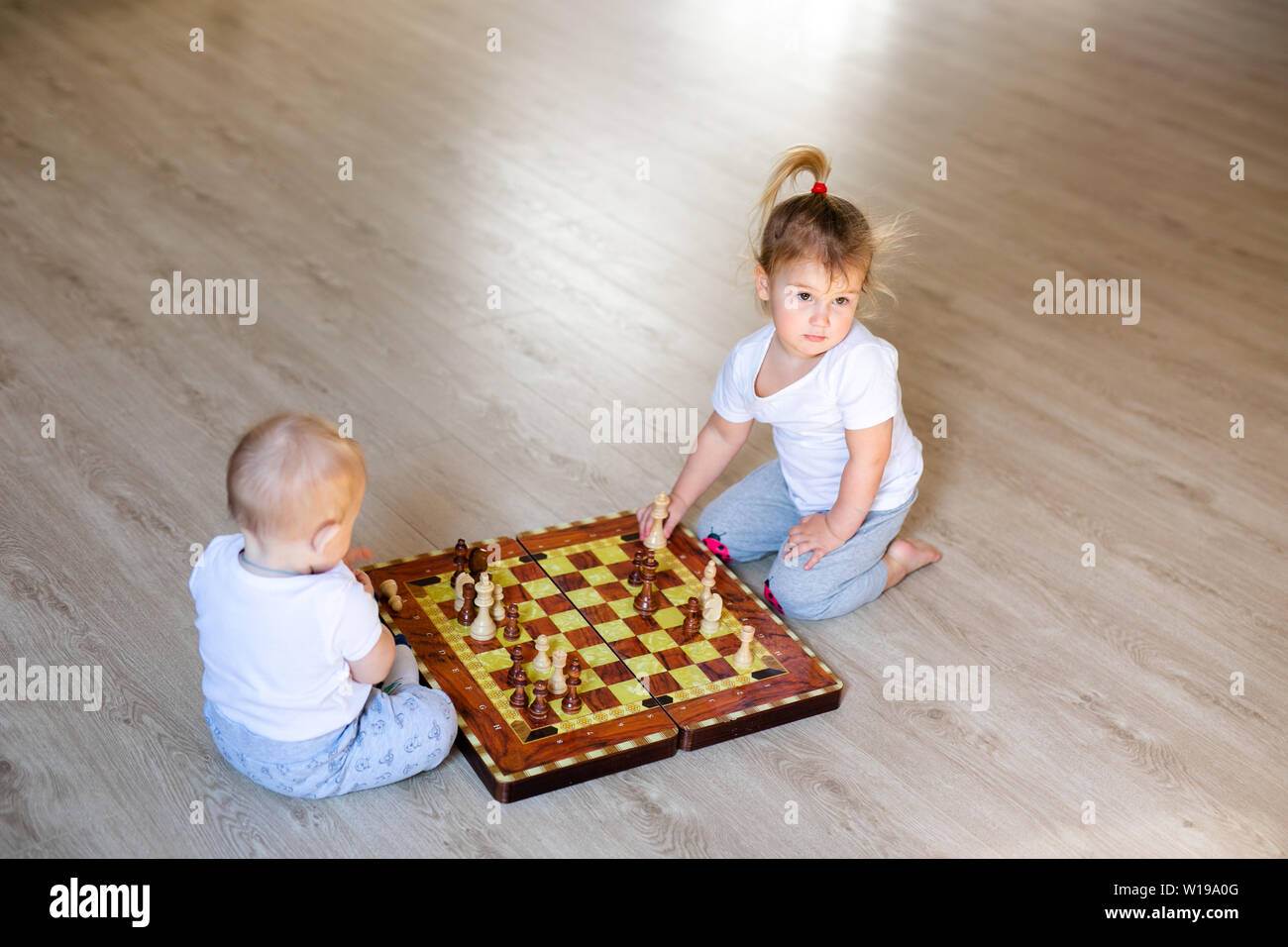 Two babies boy and girl playing chess on the white wooden floor at home ...