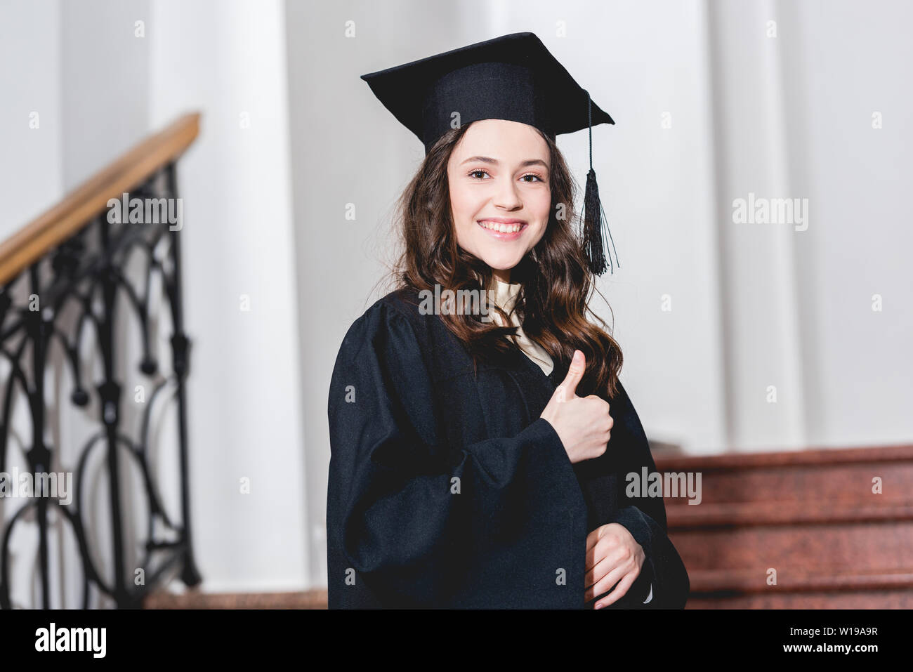 cheerful girl in graduation cap smiling while showing thumb up Stock ...