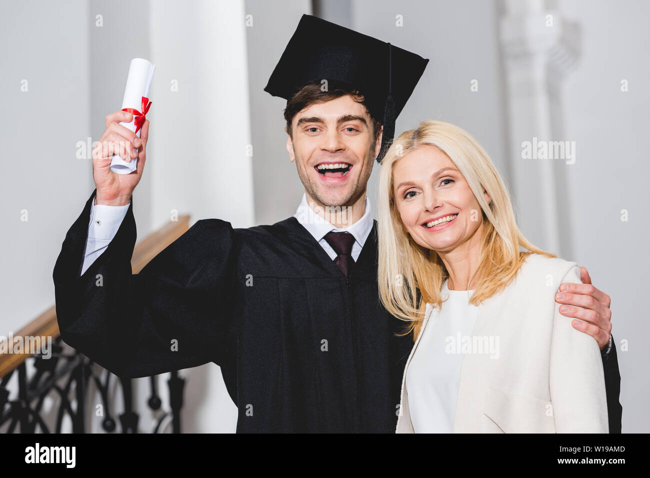 cheerful son in graduation cap holding diploma while standing with ...