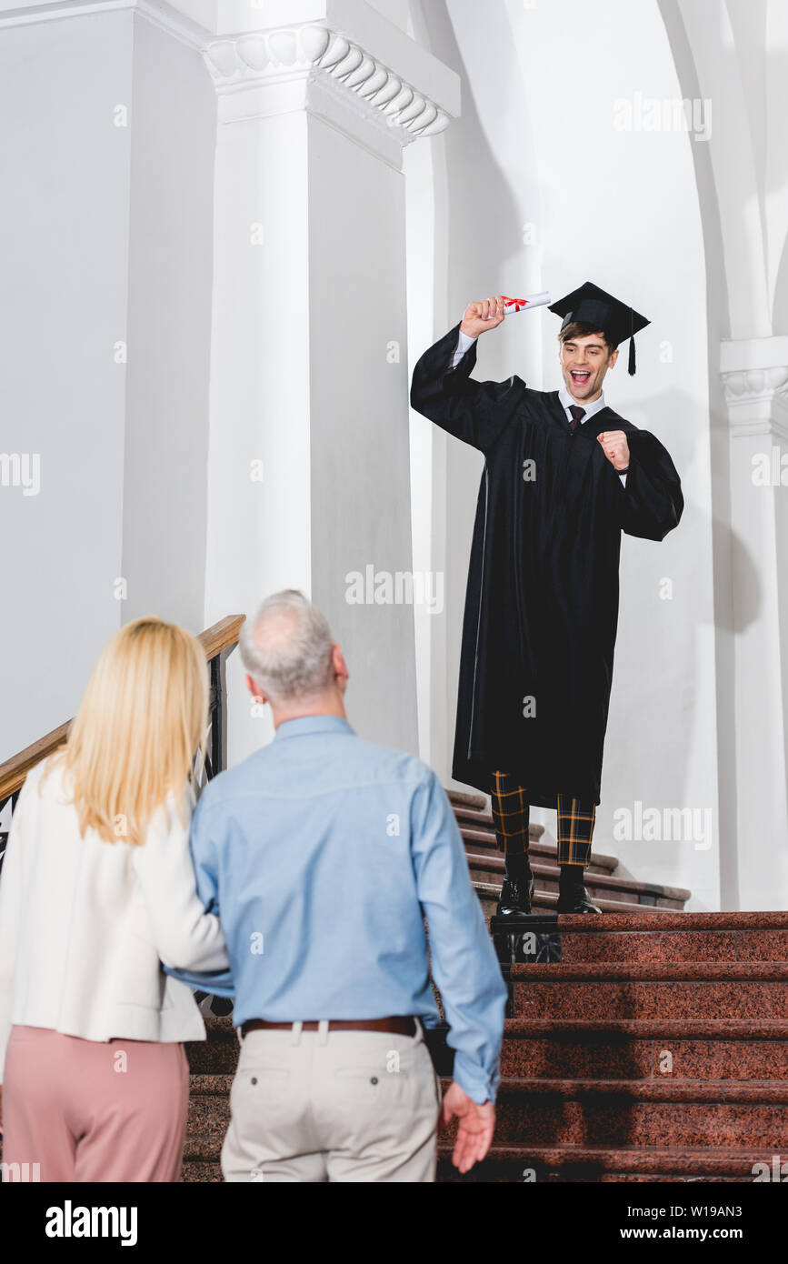 back view of parents looking at happy son in graduation cap gesturing ...