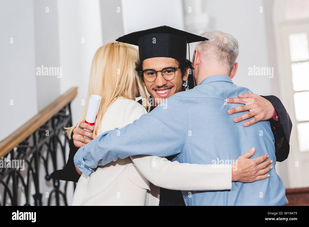 back view of mature parents hugging happy son in glasses and graduation ...