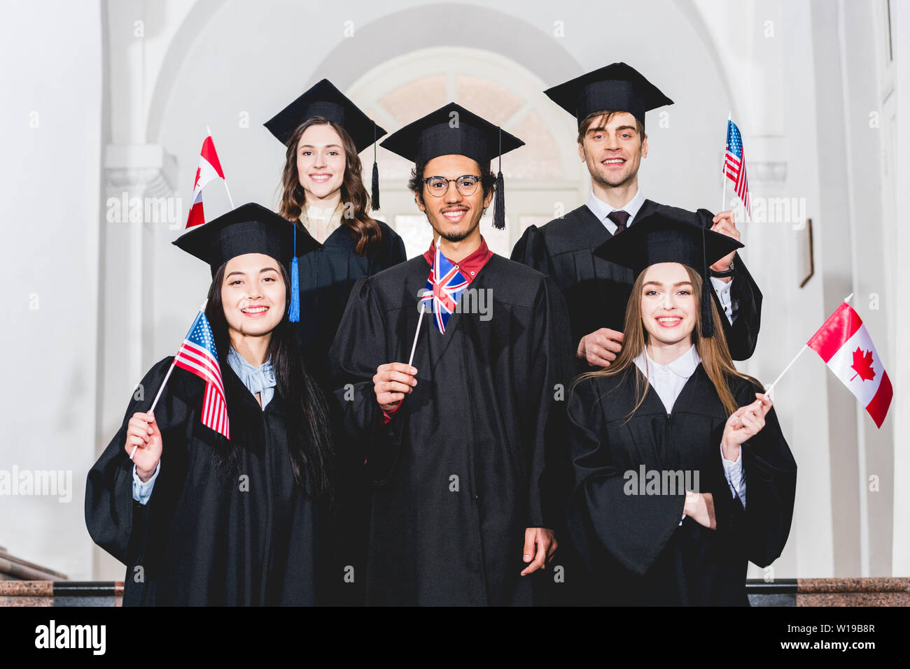 happy students in graduation gowns holding flags of different countries ...