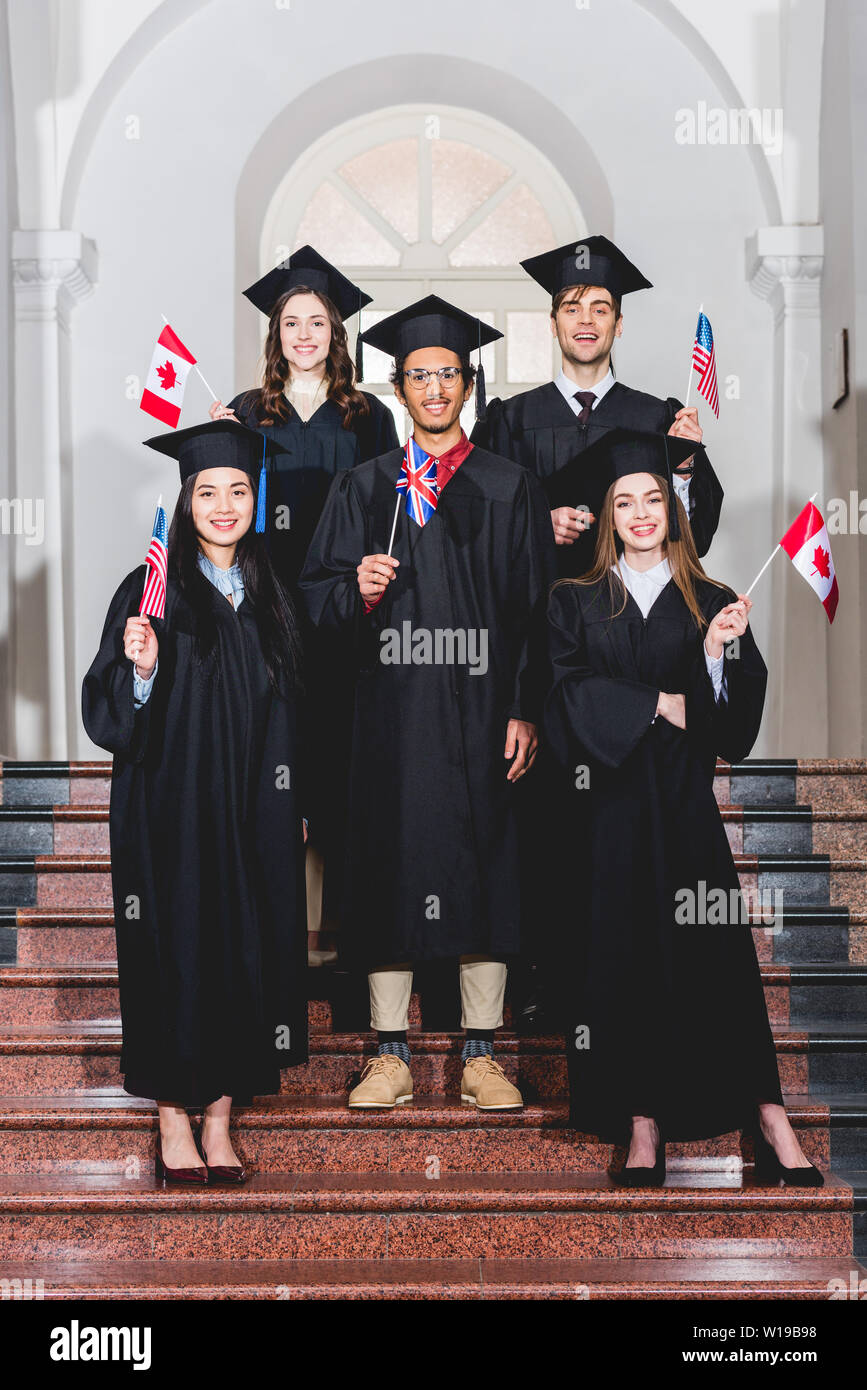 cheerful students in graduation gowns holding flags of different ...