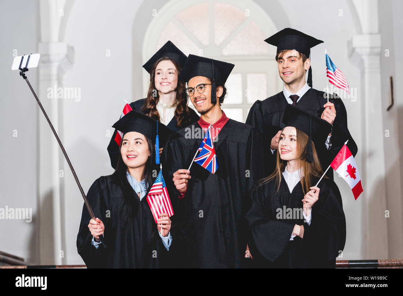 cheerful students in graduation gowns holding flags of different ...