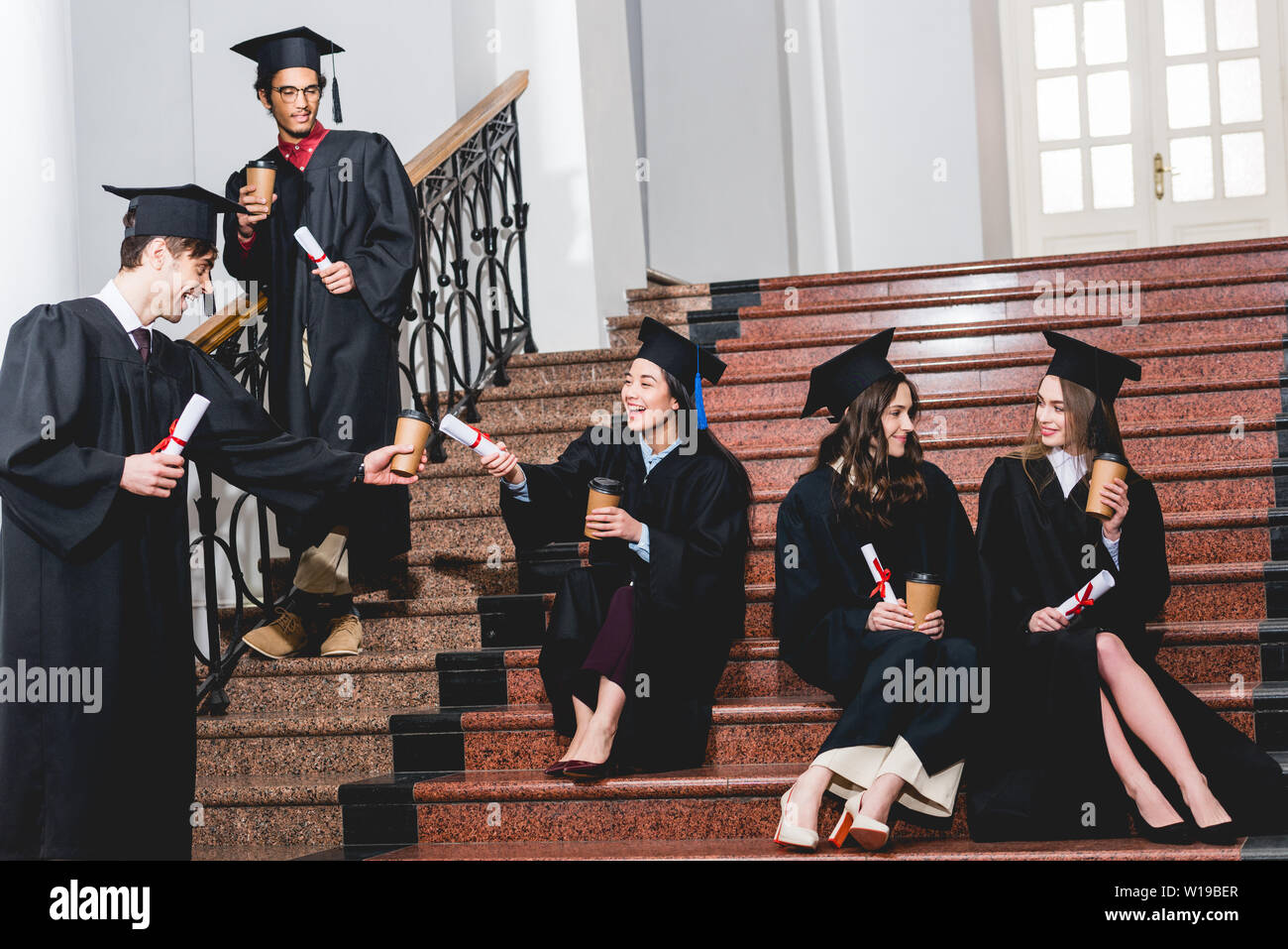 cheerful group of students in graduation gowns sitting on stairs with ...