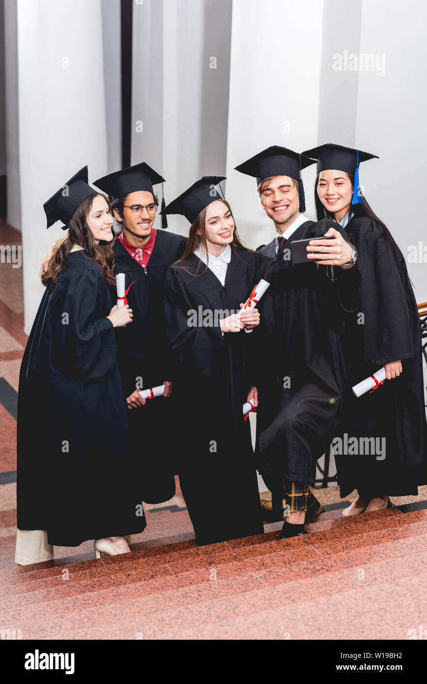 happy students in graduation gowns taking selfie and smiling while ...