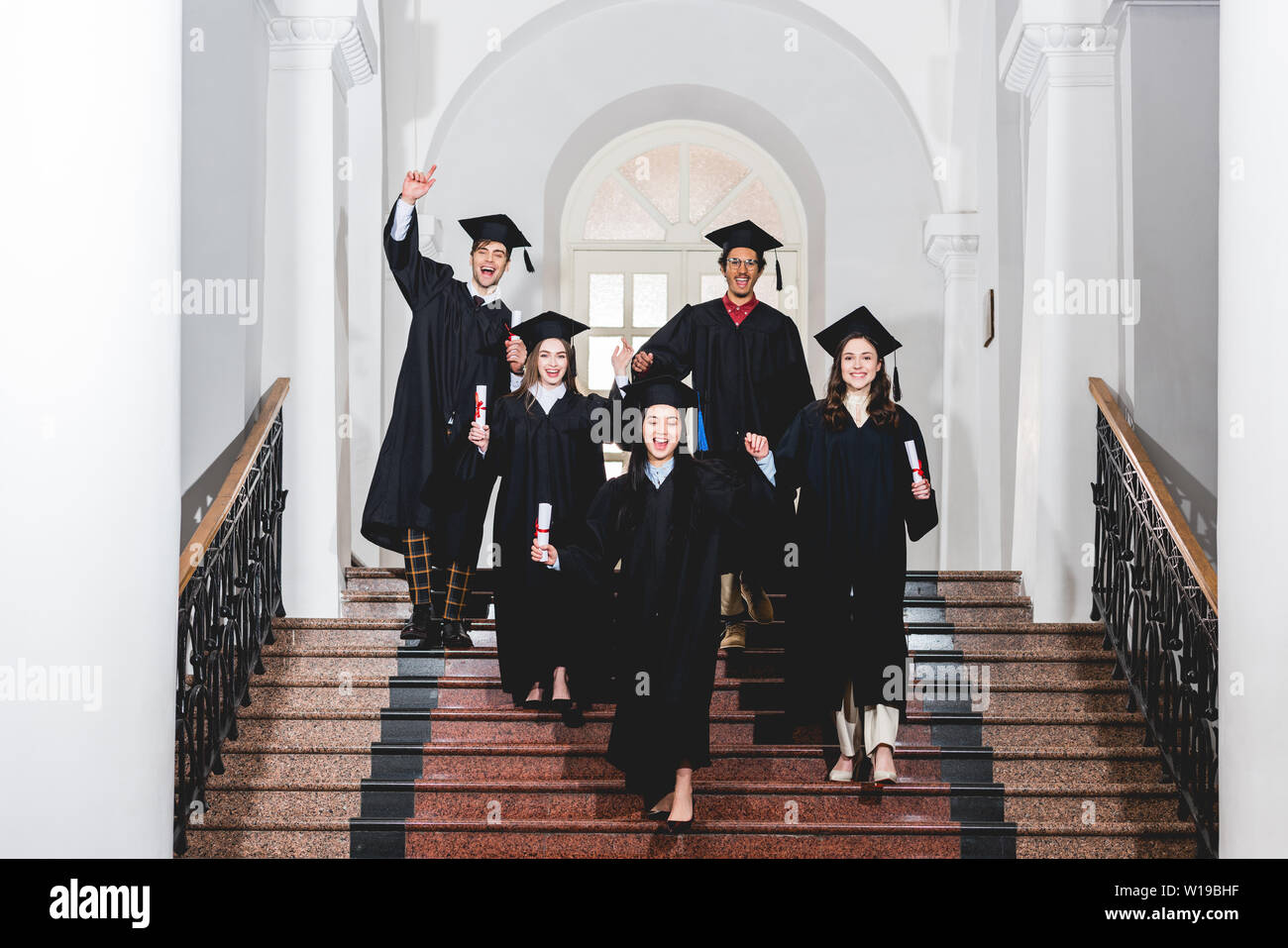 cheerful group of students in graduation gowns gesturing while holding ...