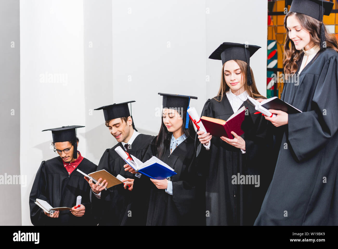 cheerful students in graduation gowns holding diplomas and reading ...