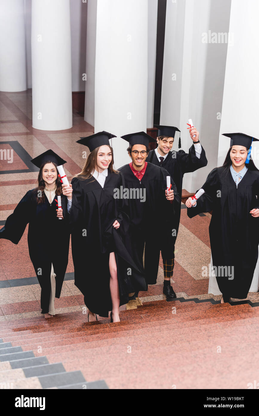 cheerful students in graduation gowns holding diplomas and walking on ...