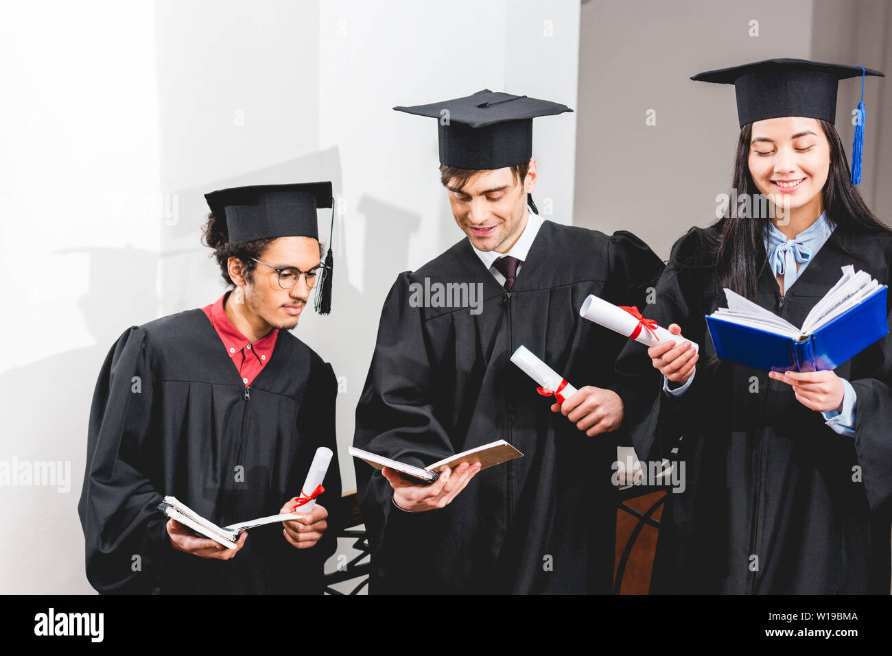 happy girl reading book near students in graduation caps holding ...