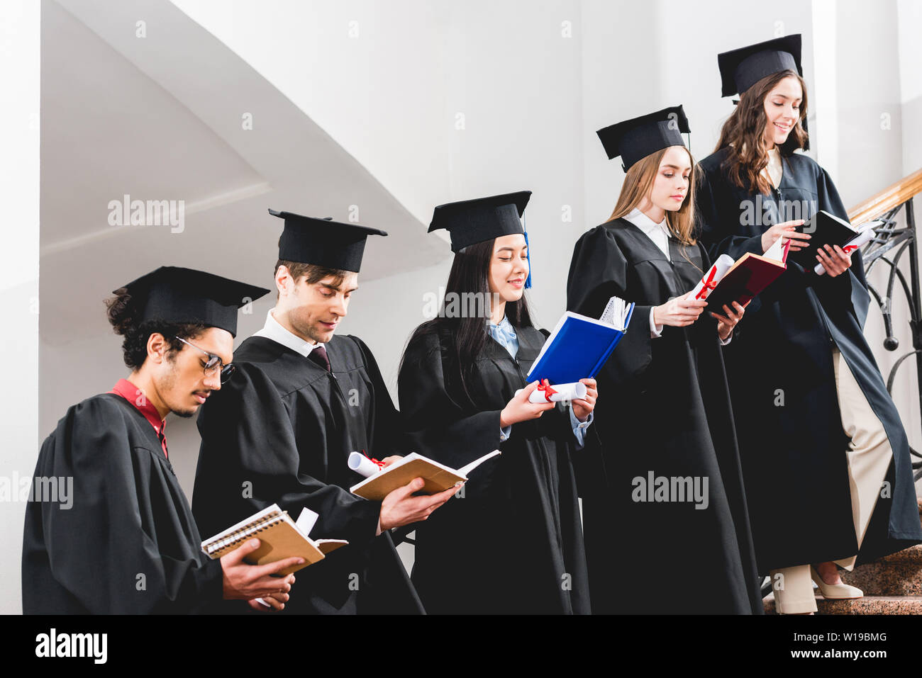 happy students in graduation gowns holding diplomas and reading books ...