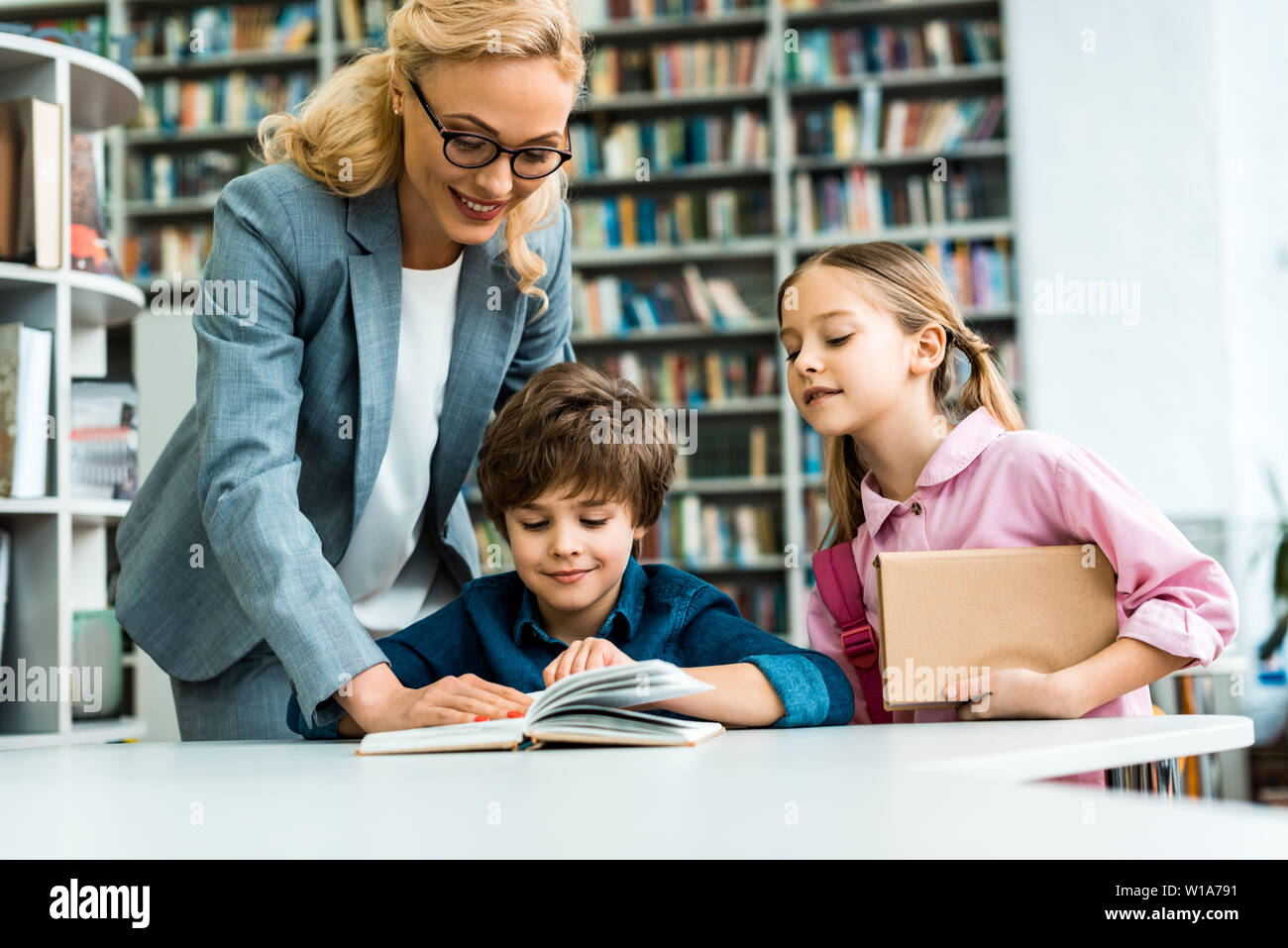 cheerful teacher in glasses standing near cute kids reading book in ...