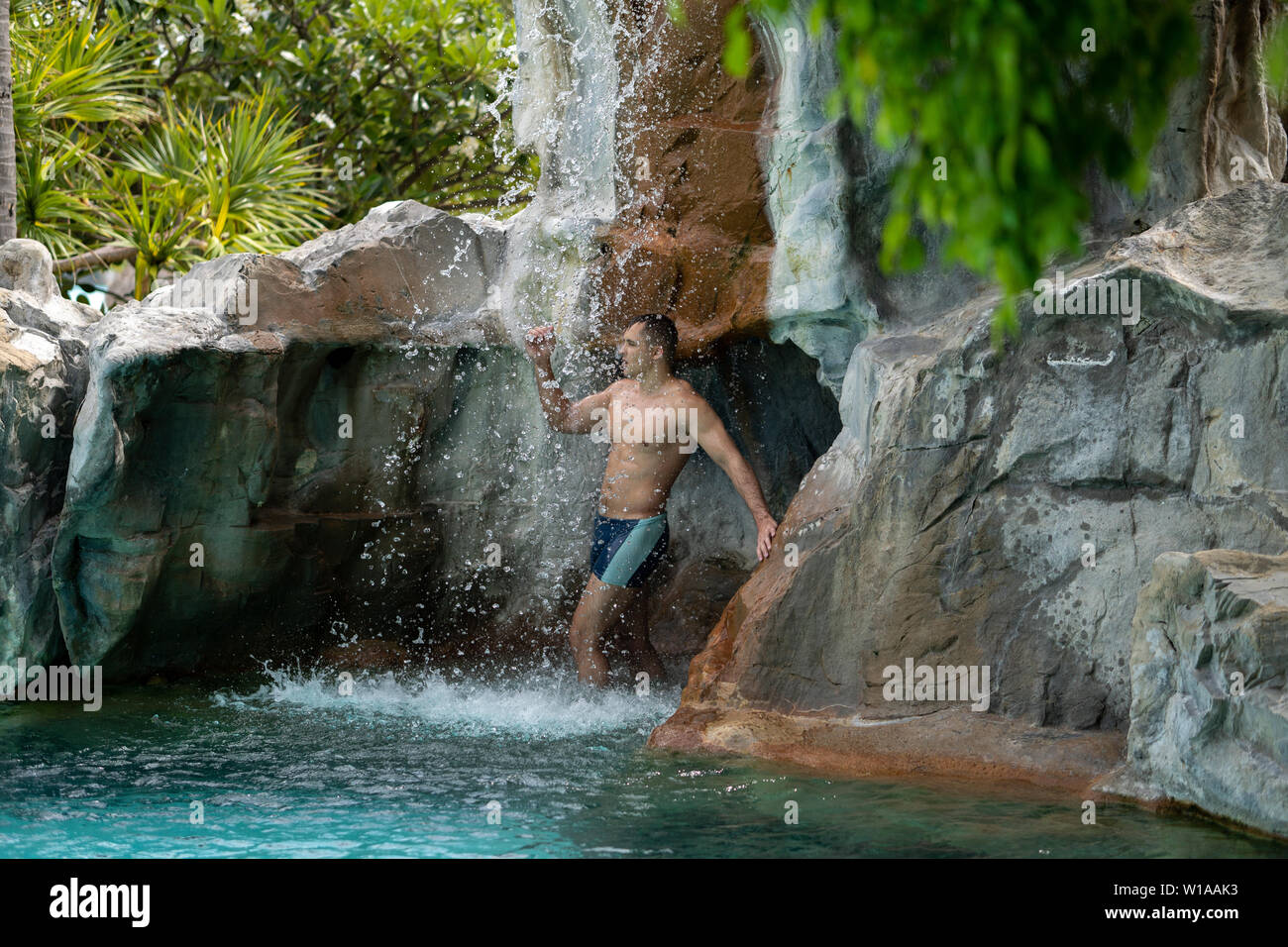 A young guy stands under artificial waterfalls in the pool on site ...