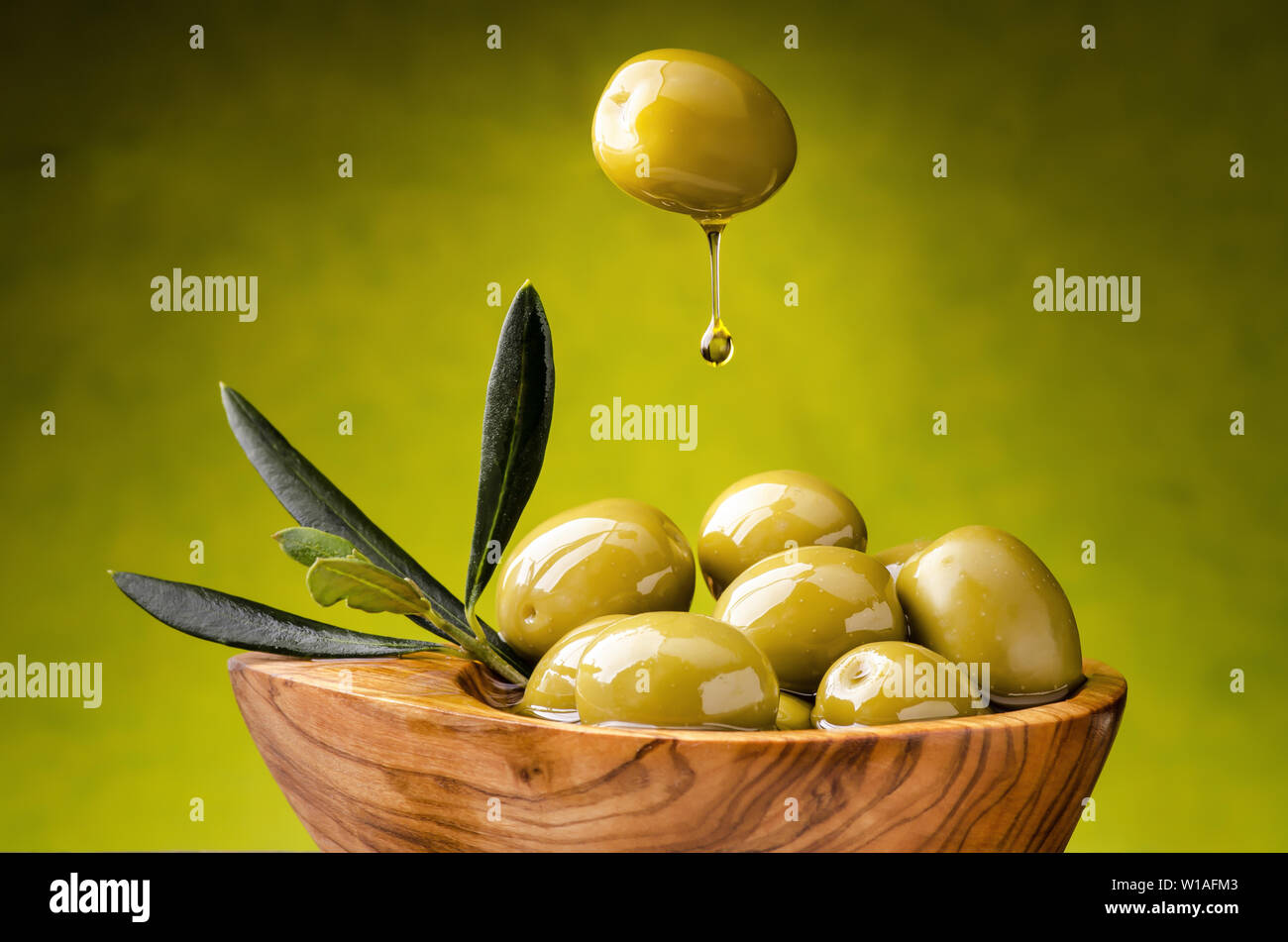 green olive with drop of olive oil falling into wooden bowl full of ...