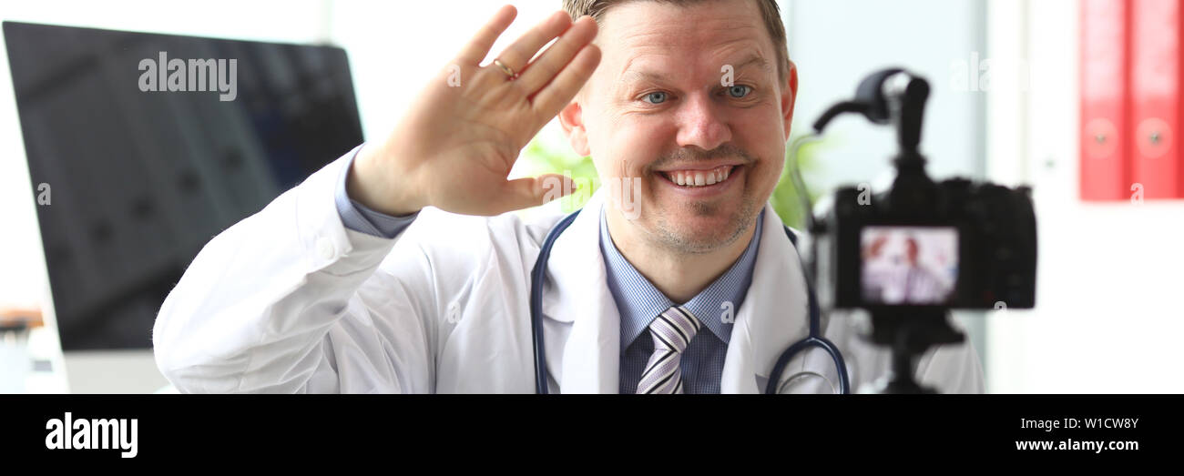 Smiling male millennial doctor wave his arm to camera saying hello to ...
