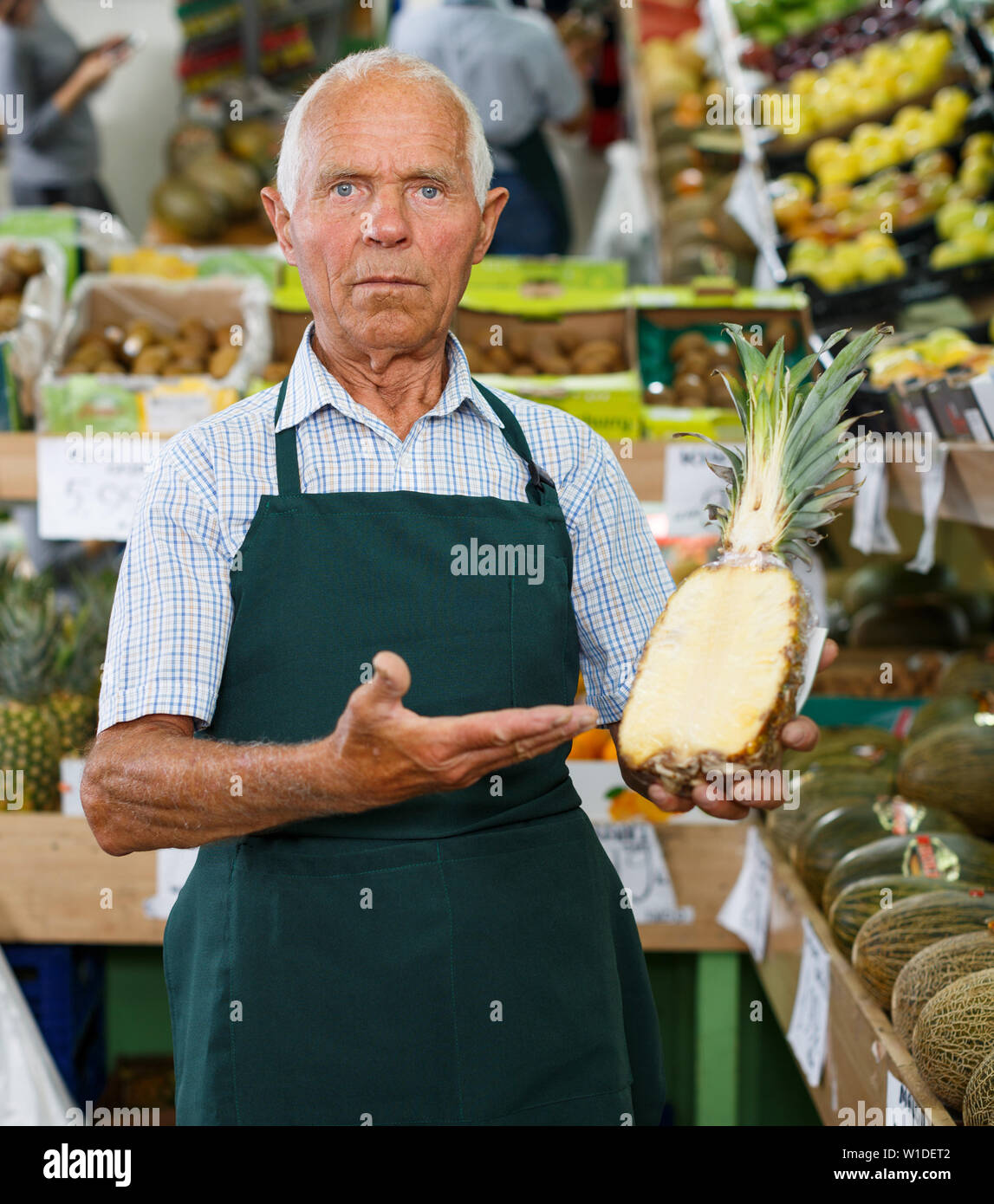 Positive senior male owner of greengrocery shop in apron offering fresh ...