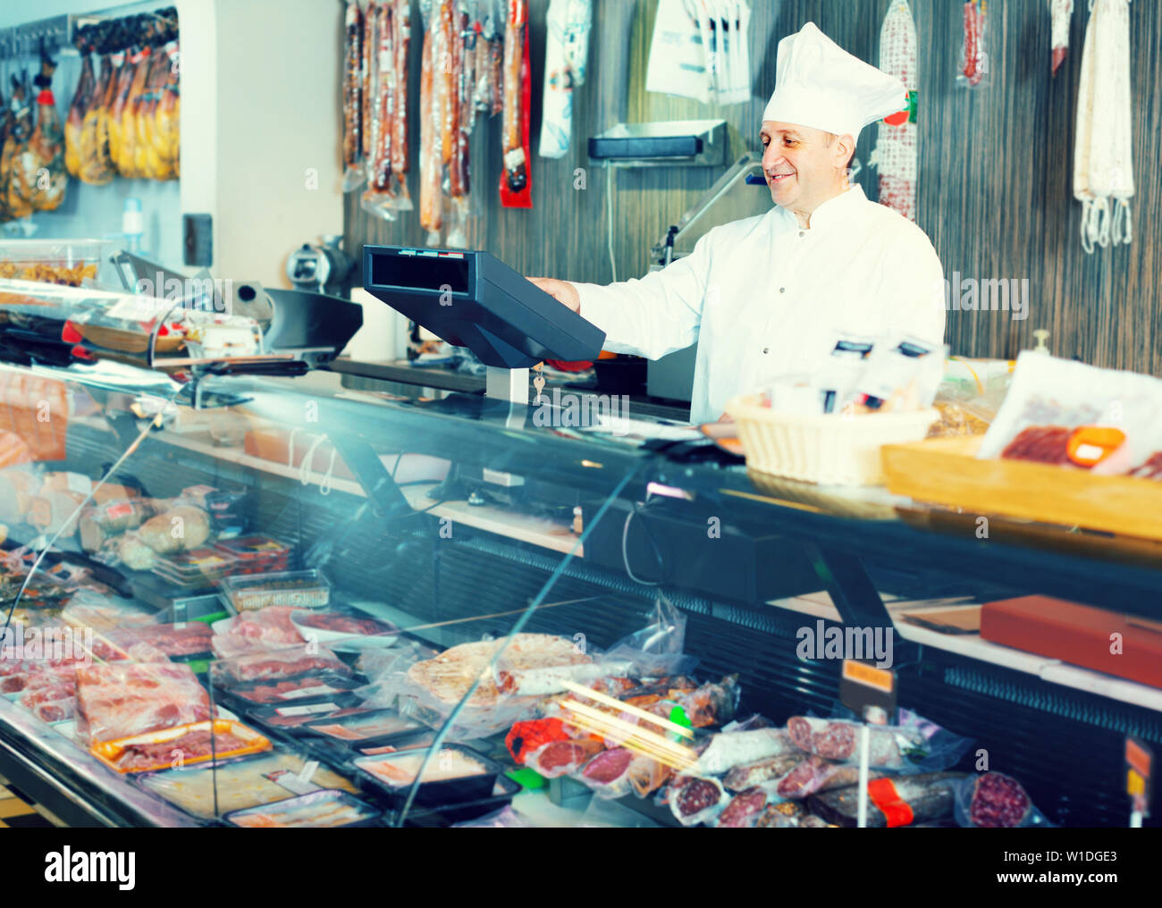 Mature male butcher with wurst, sausage and smoked meat at counter ...