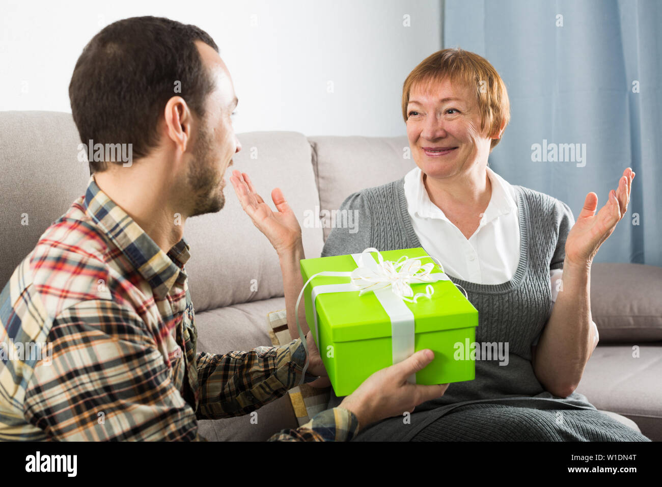 Adult son giving present to aged mother during evening together at home ...