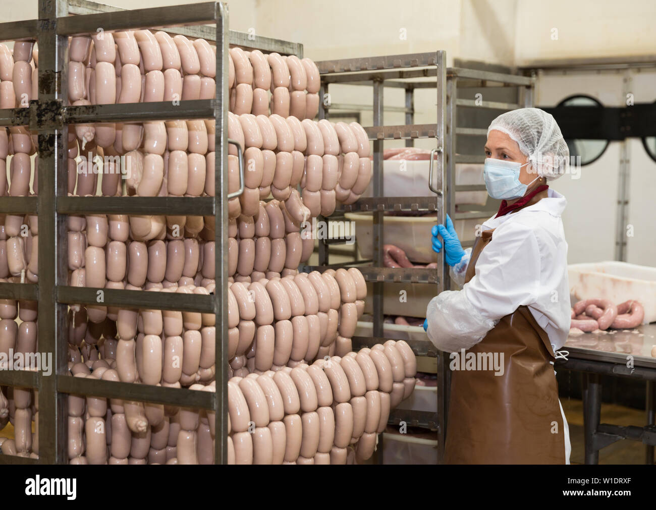 Female worker carrying raw sausages on racks in storage room at meat ...