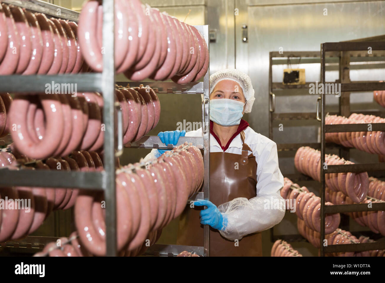 Female worker carrying raw sausages on racks in storage room at meat ...